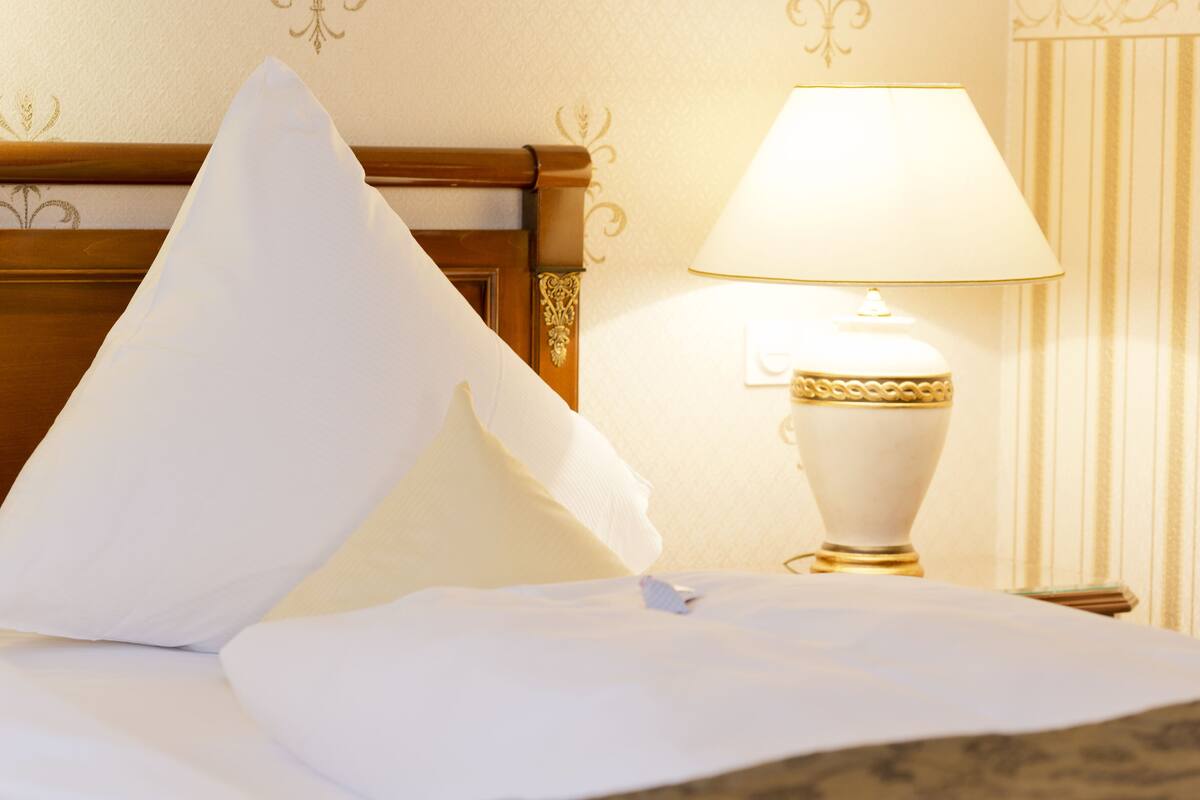 Uma cama bem arrumada com lençóis e almofadas brancos, cabeceira de madeira e abajur com cúpula branca.