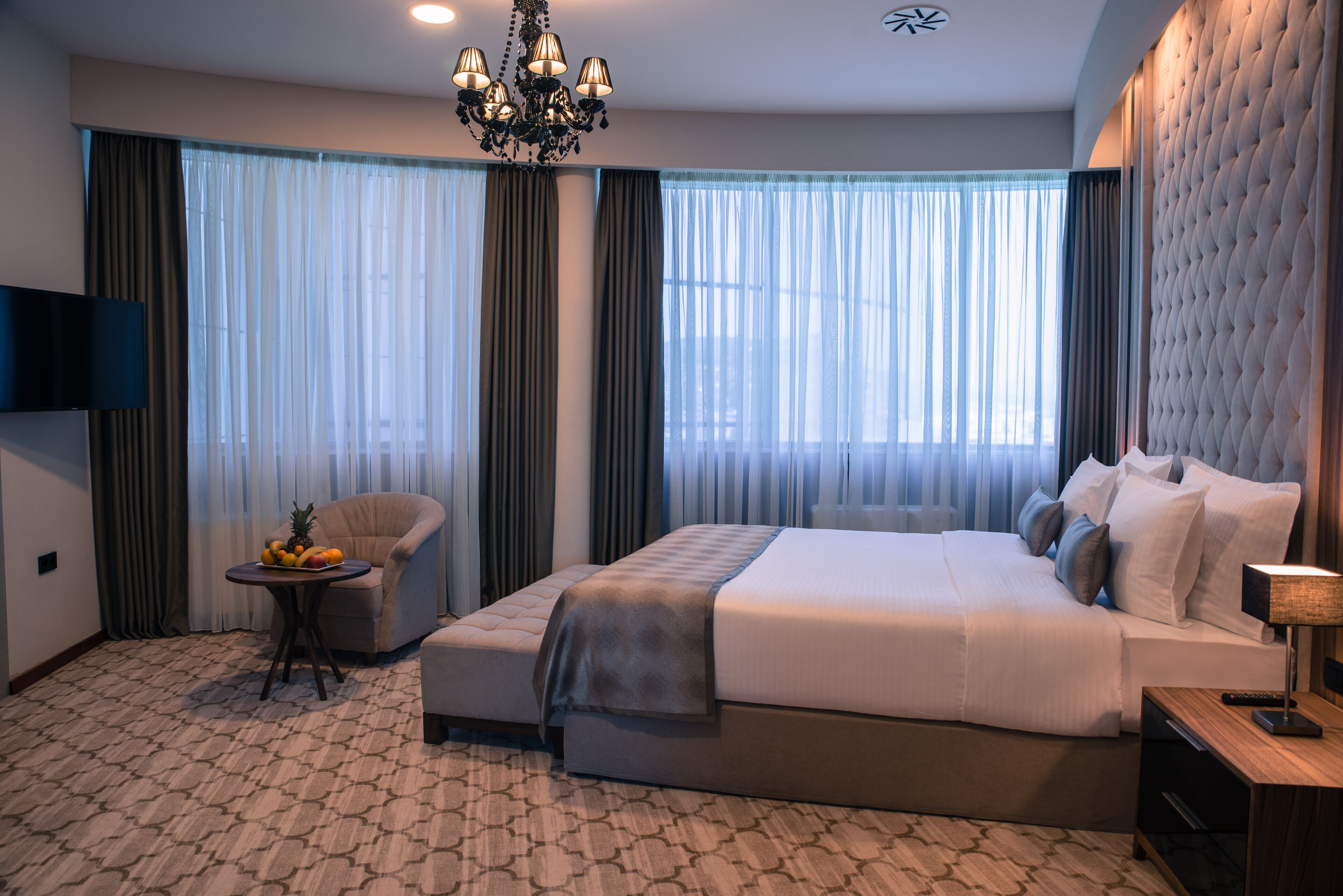 Quarto de hotel com uma cama grande, uma cadeira, uma mesa pequena com frutas e uma televisão na parede.
