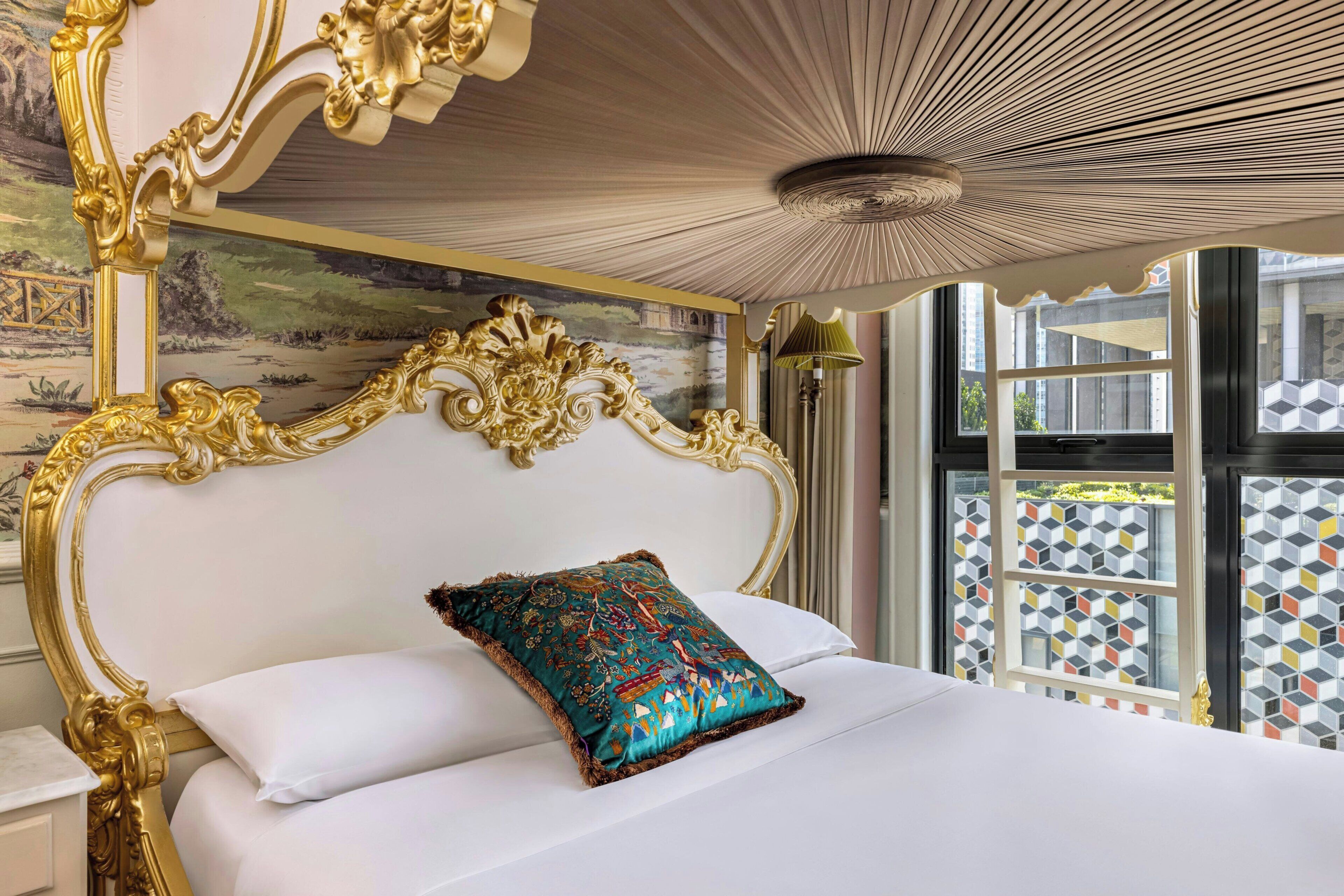 Uma cama com cabeceira branca e um travesseiro decorativo, um quadro na parede e uma janela com cortinas estampadas.