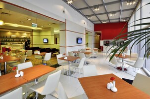 Um restaurante moderno com cadeiras brancas e mesas de madeira, uma parede de destaque vermelha e uma tela de televisão.