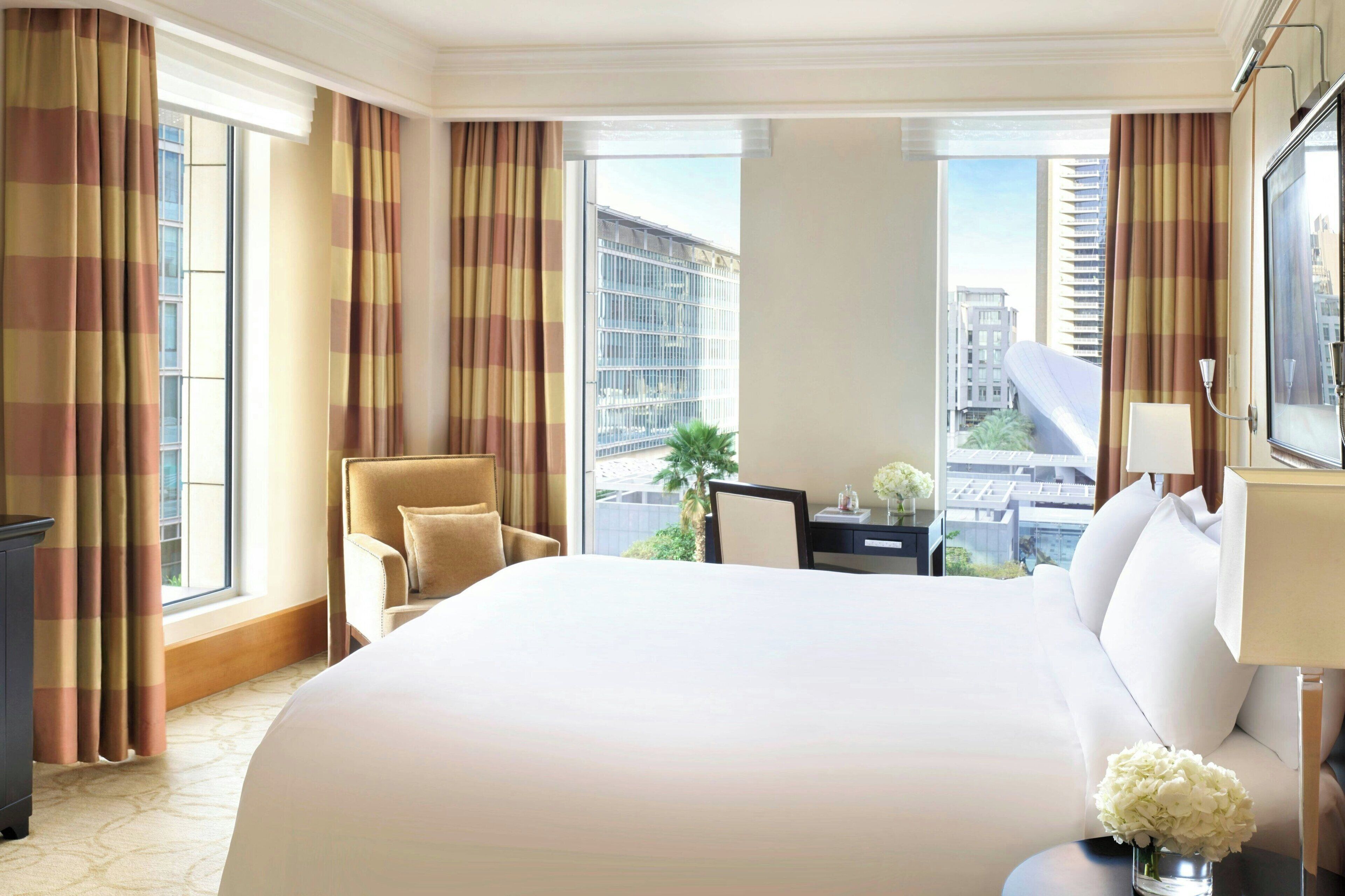 Quarto de hotel com uma cama grande, uma cadeira, uma escrivaninha com abajur e vista da cidade através de grandes janelas.