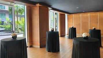 Um quarto com quatro mesas redondas, cada uma com toalha de mesa preta e um vaso de flores. O quarto possui grandes janelas com vista para palmeiras e um edifício lá fora.