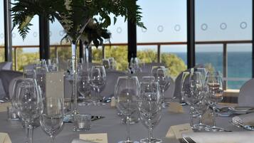 Mesa posta para um evento formal, com copos de vidro transparente, um arranjo floral e vista para o exterior.