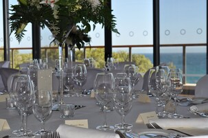 Mesa posta para um evento formal, com copos de vidro transparente, um arranjo floral e vista para o exterior.