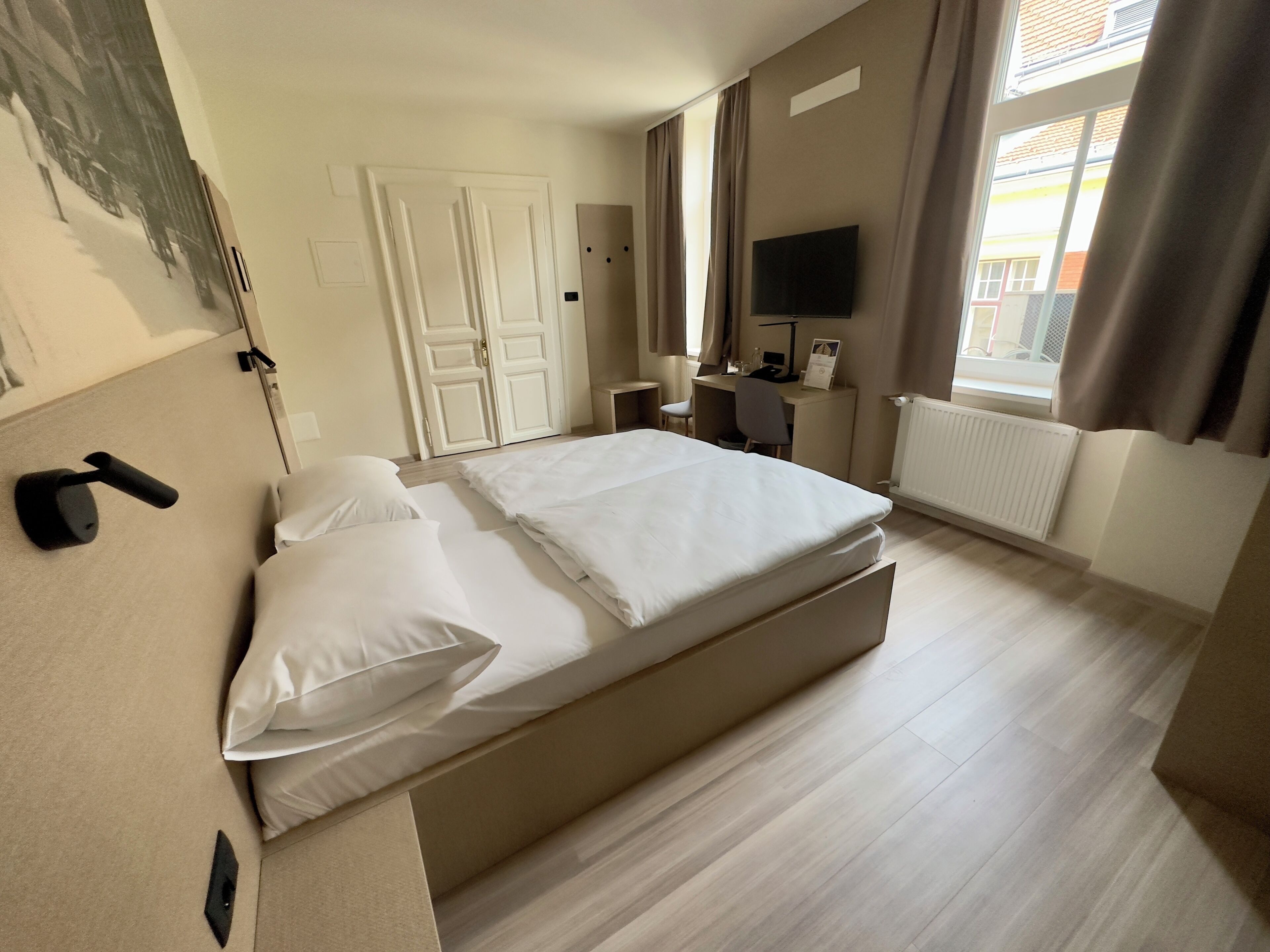 Quarto de hotel com cama, escrivaninha, cadeira, televisão e janela com cortinas.
