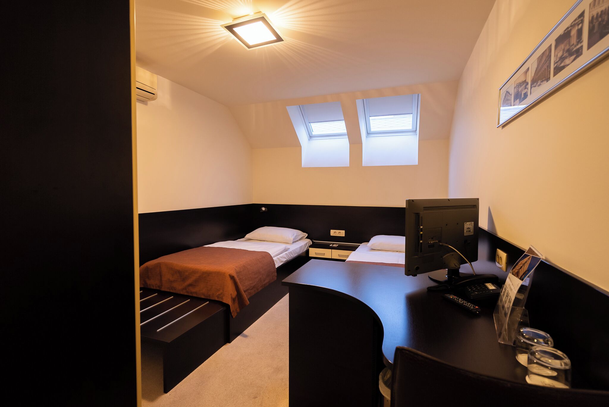 Quarto de hotel pequeno com duas camas, uma mesa, um computador e uma janela.