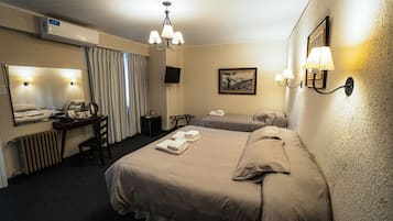Quarto de hotel com duas camas, uma mesa, uma cadeira, um espelho, um radiador, uma televisão e luminárias fixadas na parede.