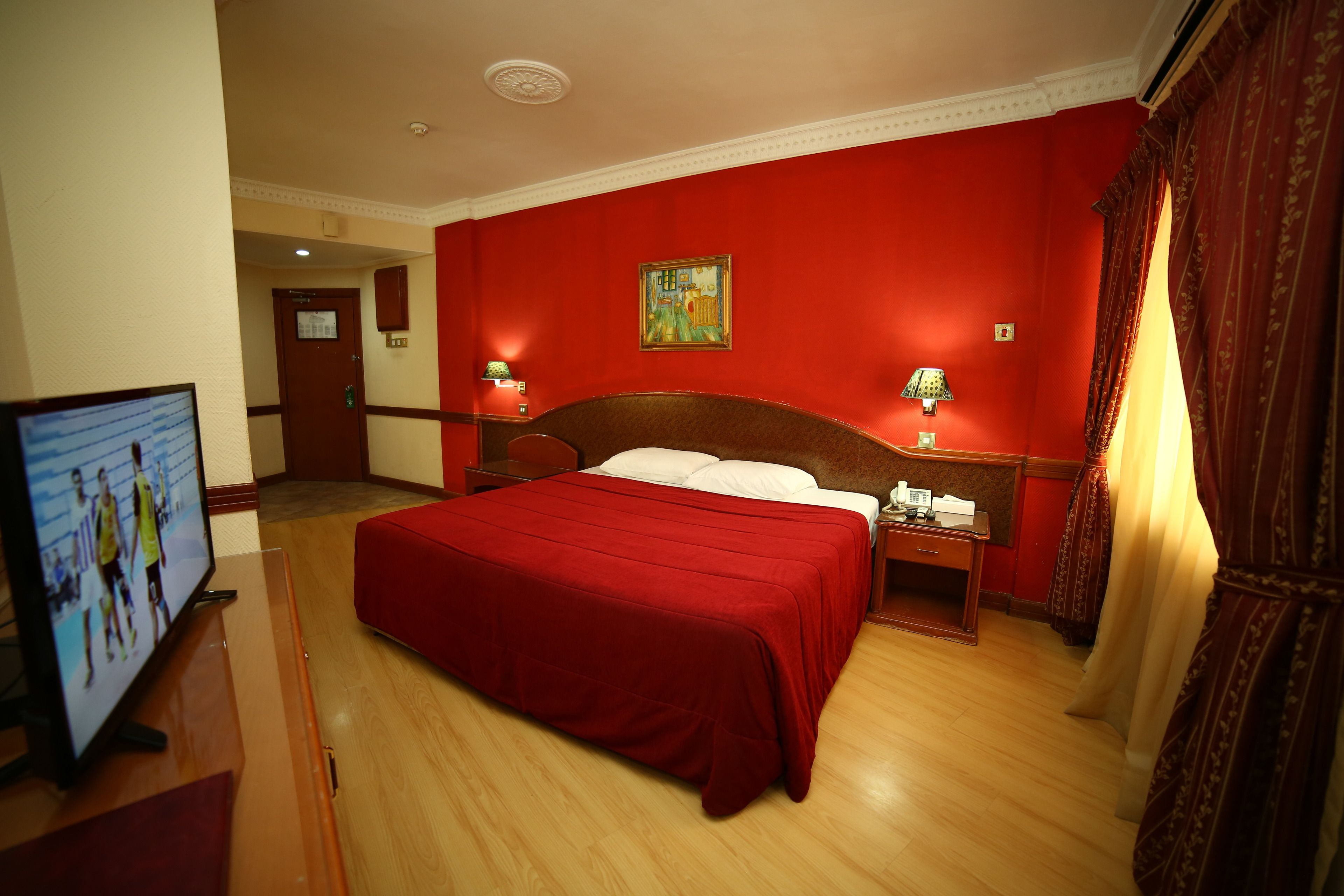 Quarto de hotel com uma cama grande, duas mesas de cabeceira com luminárias, uma televisão e uma parede vermelha com um quadro.