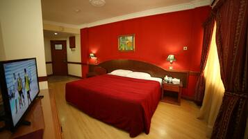 Quarto de hotel com uma cama grande, duas mesas de cabeceira com luminárias, uma televisão e uma parede vermelha com um quadro.