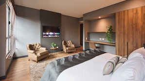 Quarto moderno com uma cama grande, duas poltronas de couro, uma mesinha com uma planta, uma televisão montada e uma estante de madeira com uma planta.