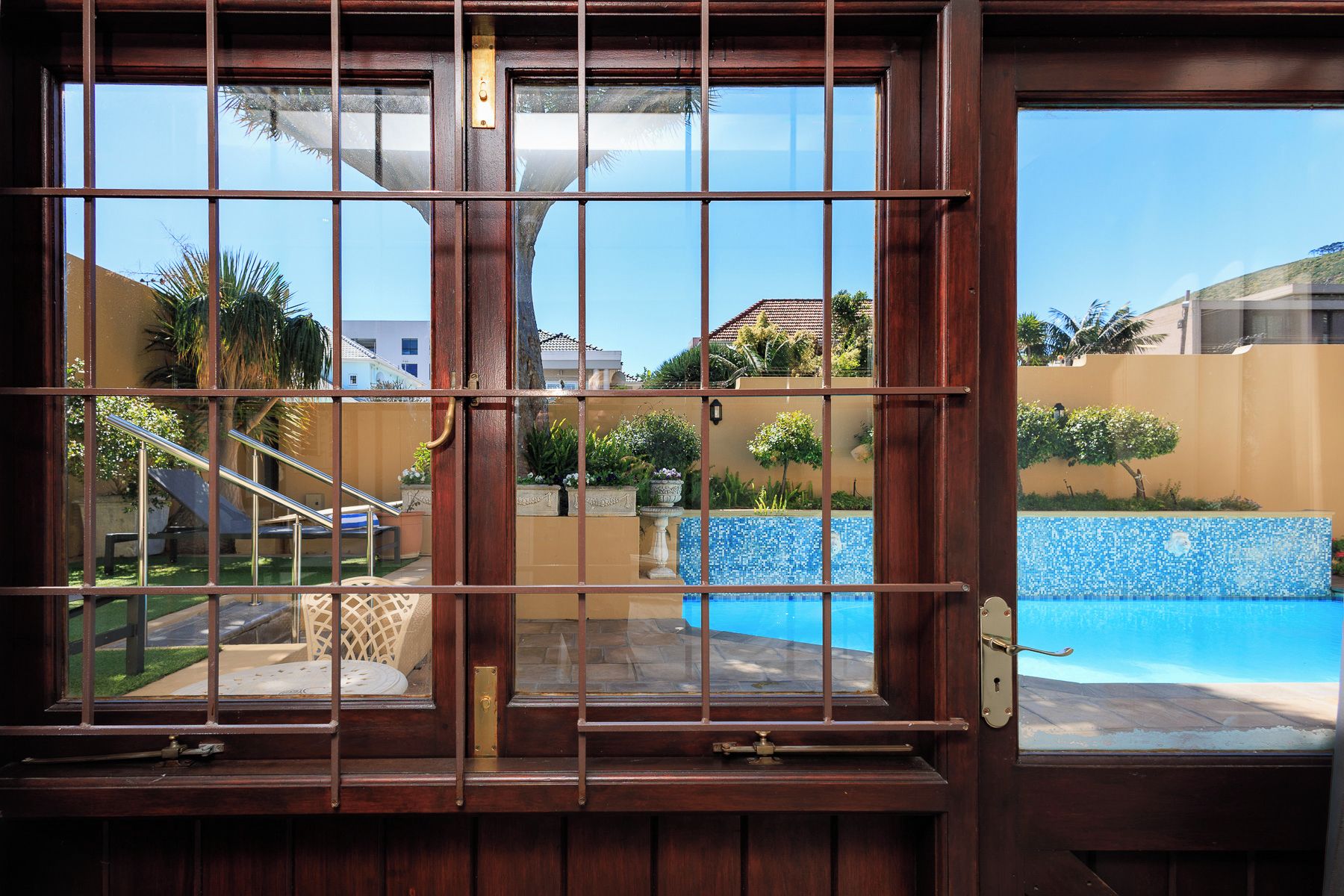 Vista através de uma janela de madeira com grades de metal, com piscina e área ajardinada com plantas e escada.