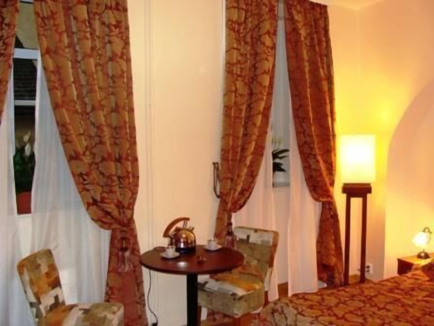 Quarto de hotel com uma cama, uma mesinha redonda com chaleira, duas cadeiras, um abajur e cortinas.
