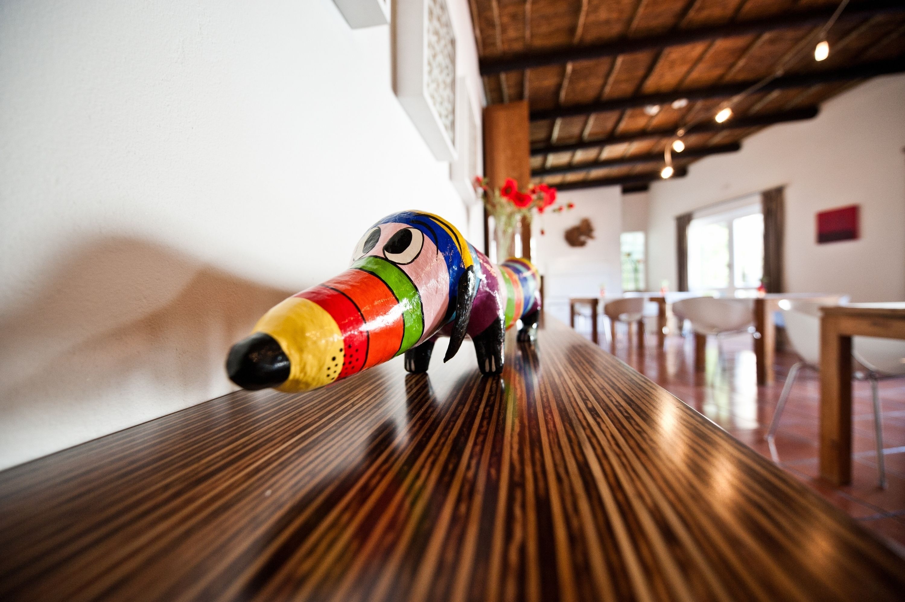 Uma escultura abstrata e colorida sobre uma mesa de madeira, num espaço com teto de madeira e paredes brancas.