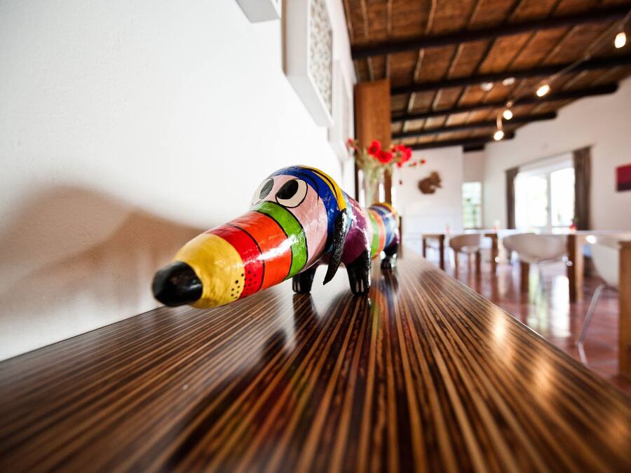 Uma escultura abstrata e colorida sobre uma mesa de madeira, num espaço com teto de madeira e paredes brancas.