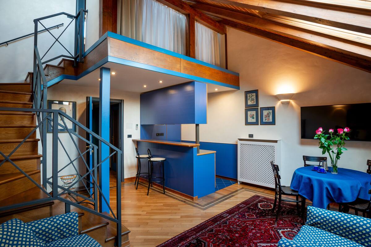 Quarto moderno estilo loft, com escada de madeira, parede azul decorativa, área de refeições com mesa e cadeiras, e uma grande janela com cortinas.