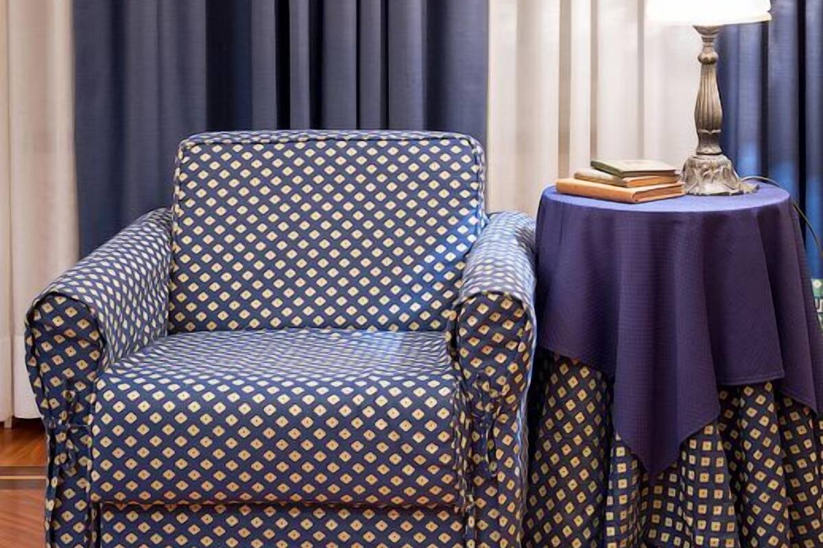 Quarto com poltrona estampada, mesa redonda pequena com abajur e vista para cortinas verticais.