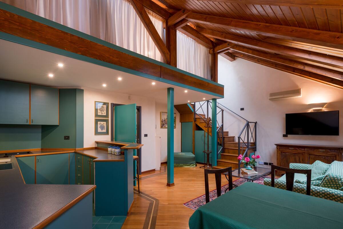 Um amplo loft com pé direito alto, vigas de madeira, área de cozinha, escada e um espaço de estar com televisão.