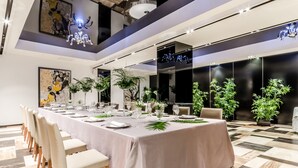 Uma longa mesa de jantar posta com cadeiras, cercada por plantas em vasos, em um interior moderno com um lustre e obras de arte nas paredes.