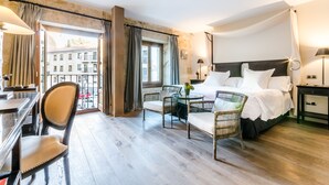 Quarto de hotel com uma cama grande, duas cadeiras de vime, uma mesa pequena e vista da cidade através de uma janela ampla.