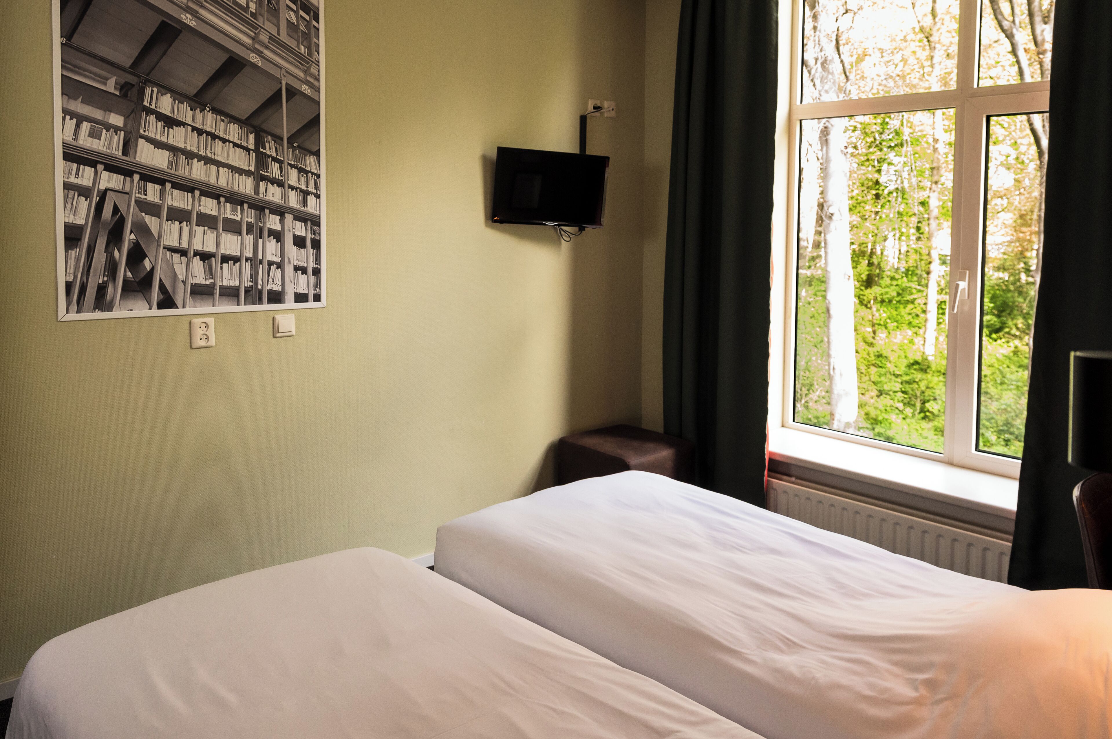 Quarto de hotel com cama, estante, televisão e janela com vista para árvores.