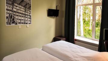 Quarto de hotel com cama, estante, televisão e janela com vista para árvores.