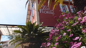 Hotel chamado "Villa Florida" com fachada vermelha, palmeiras e flores cor-de-rosa ao primeiro plano.
