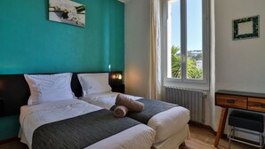 Uma cama de casal com lençóis brancos e um cobertor cinza escuro, uma escrivaninha pequena com uma cadeira, uma janela com vista para o verde e um quadro na parede.