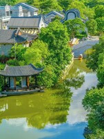 Uma cidade tradicional chinesa às margens do rio, com um pavilhão, pontes de pedra e vegetação exuberante.