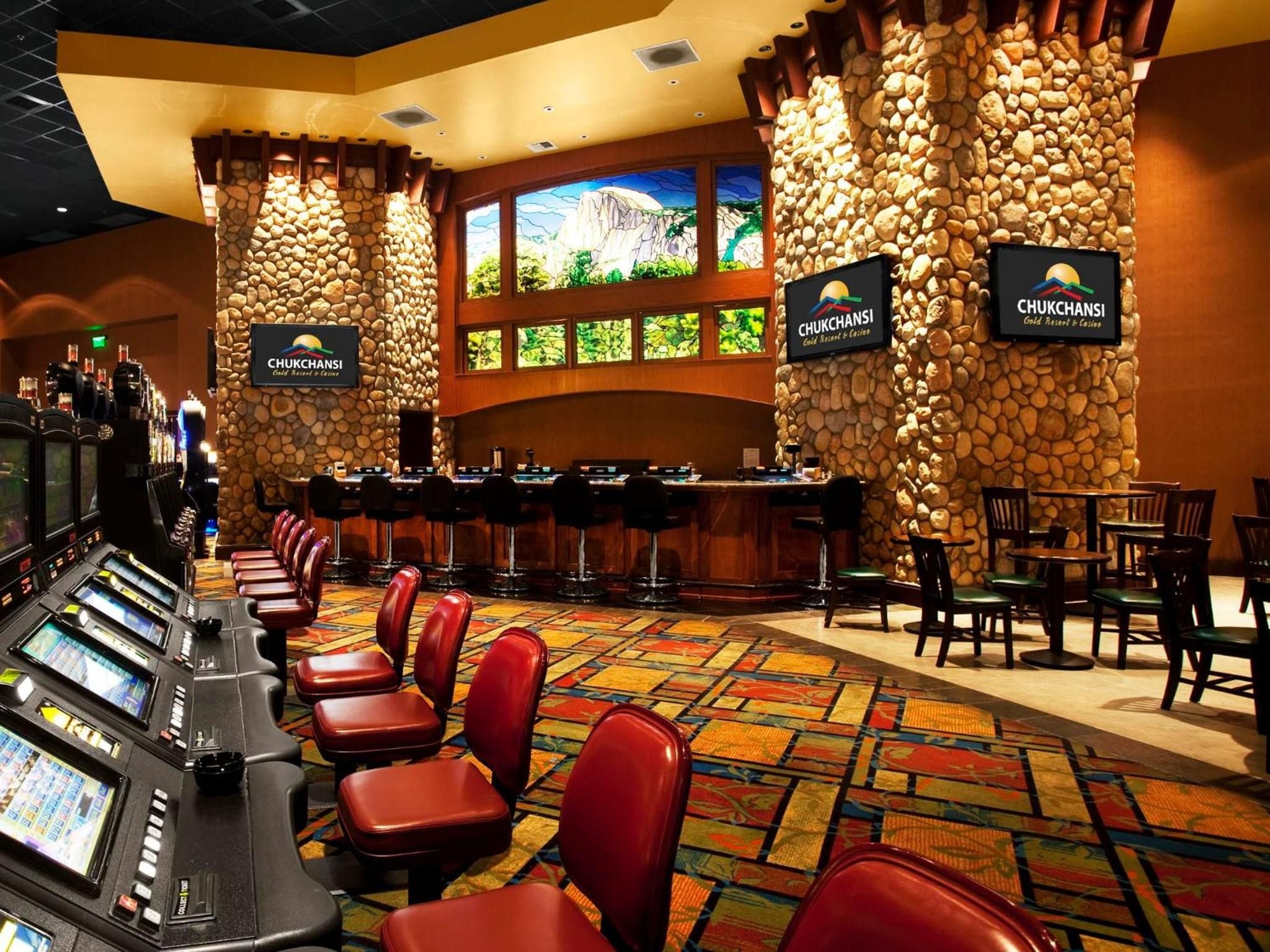 Área de casino com máquinas de slot, cadeiras vermelhas e bar com parede de pedra ao fundo.