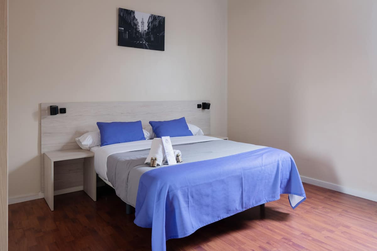 Uma cama bem arrumada com colcha azul e almofadas, uma mesa de cabeceira com abajur, e um quadro pendurado na parede.