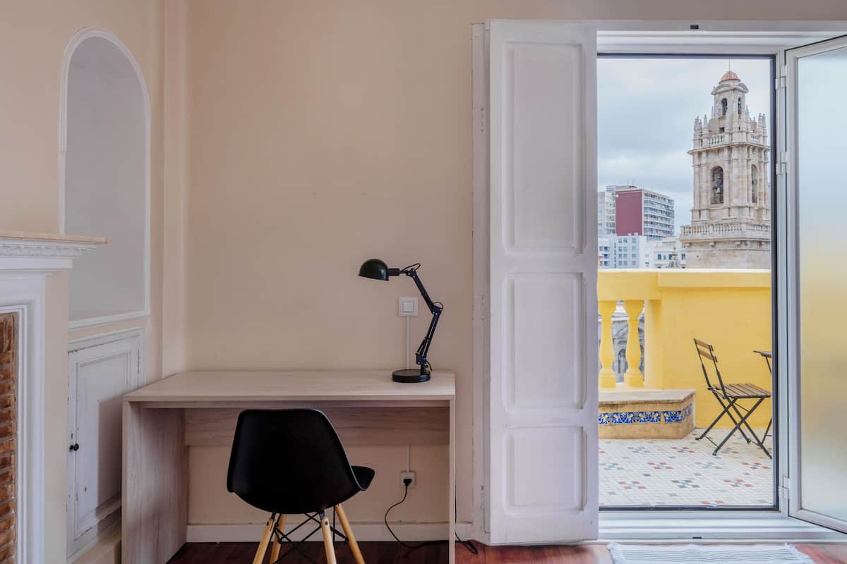 Quarto com escrivaninha, cadeira e vista para uma torre histórica através de uma porta aberta.