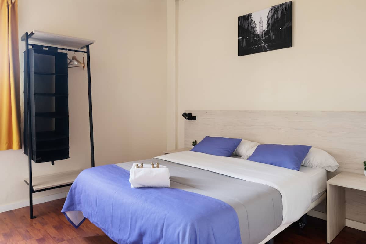 Uma cama bem arrumada com um edredom azul e branco, uma mesa de cabeceira de madeira e um suporte de cabides preto em frente a uma parede com um quadro emoldurado.
