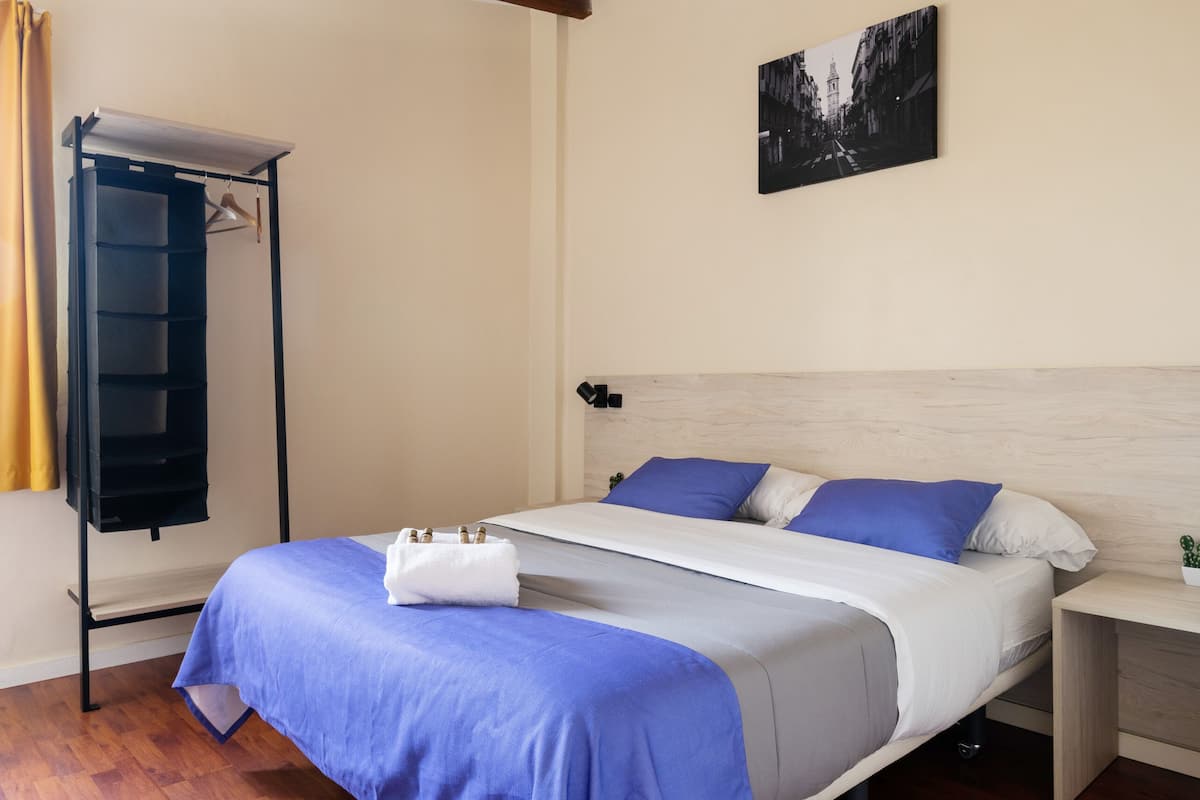 Quarto com cama, mesa de cabeceira, prateleira na parede e janela com cortinas.