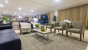 Um saguão de hotel com balcão de recepção, área de estar com sofás e poltronas, e uma grande janela com cortinas.