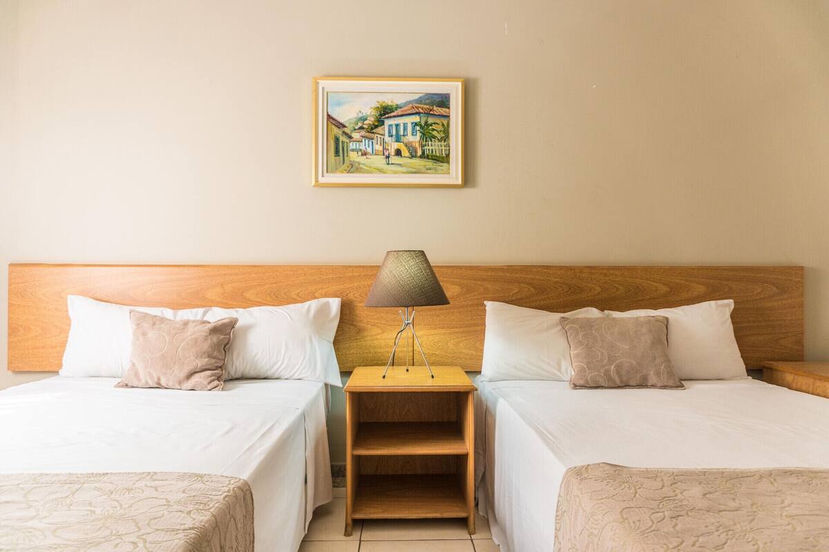 Quarto de hotel com duas camas, uma mesinha de cabeceira, uma luminária e um quadro na parede.