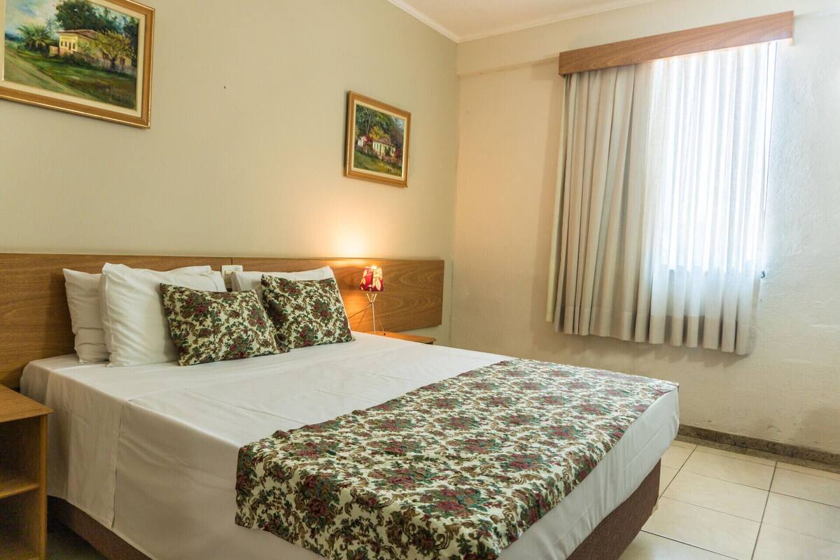 Quarto de hotel com cama, mesa de cabeceira, luminária, janela com cortinas e duas pinturas emolduradas na parede.