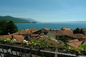 Uma vila costeira com casas de telhados em terracota, vista clara do mar e montanhas ao fundo.