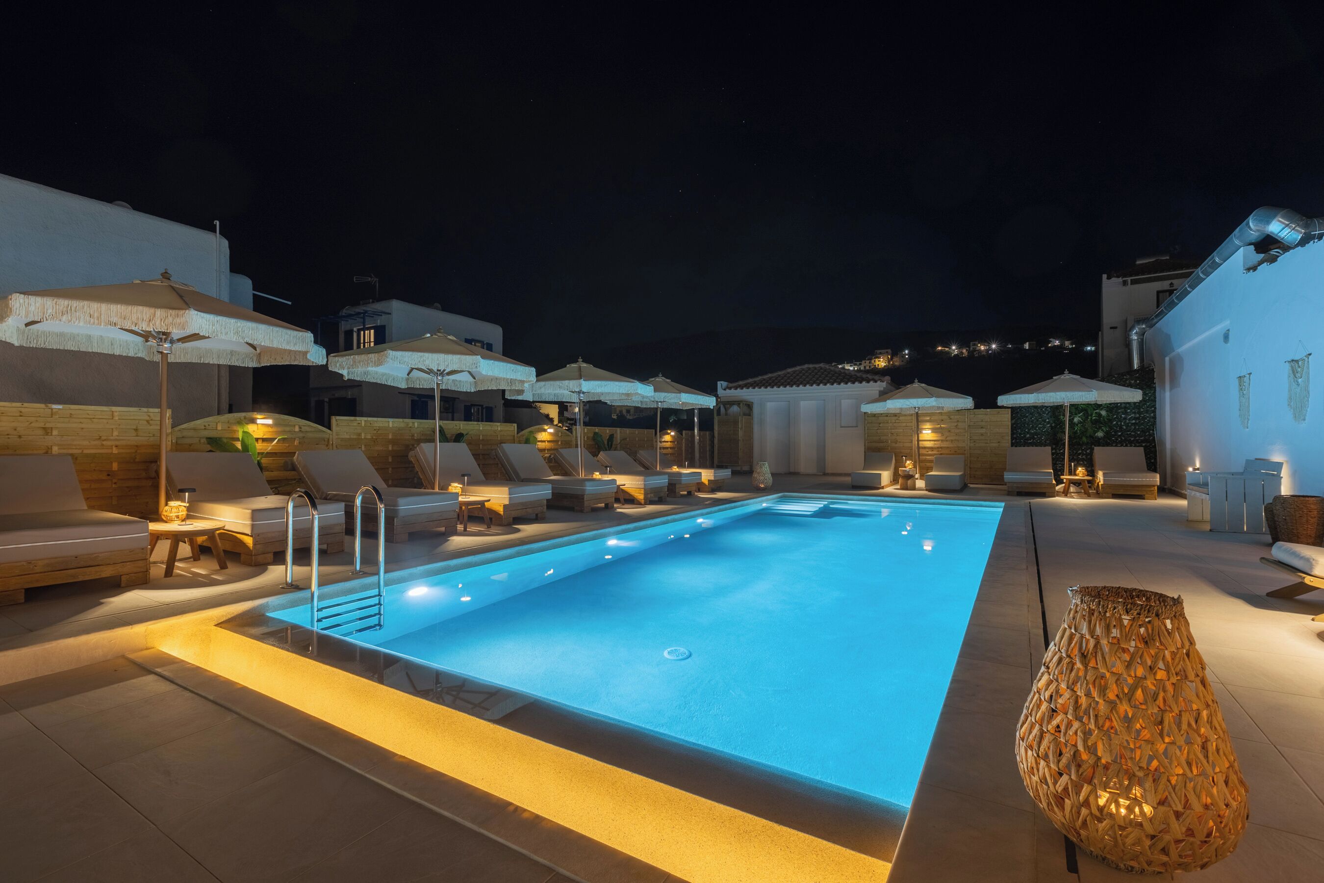 Uma área de piscina à noite, com espreguiçadeiras e guarda-sóis, cercada por edifícios.