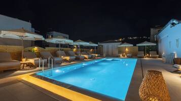 Uma área de piscina à noite, com espreguiçadeiras e guarda-sóis, cercada por edifícios.