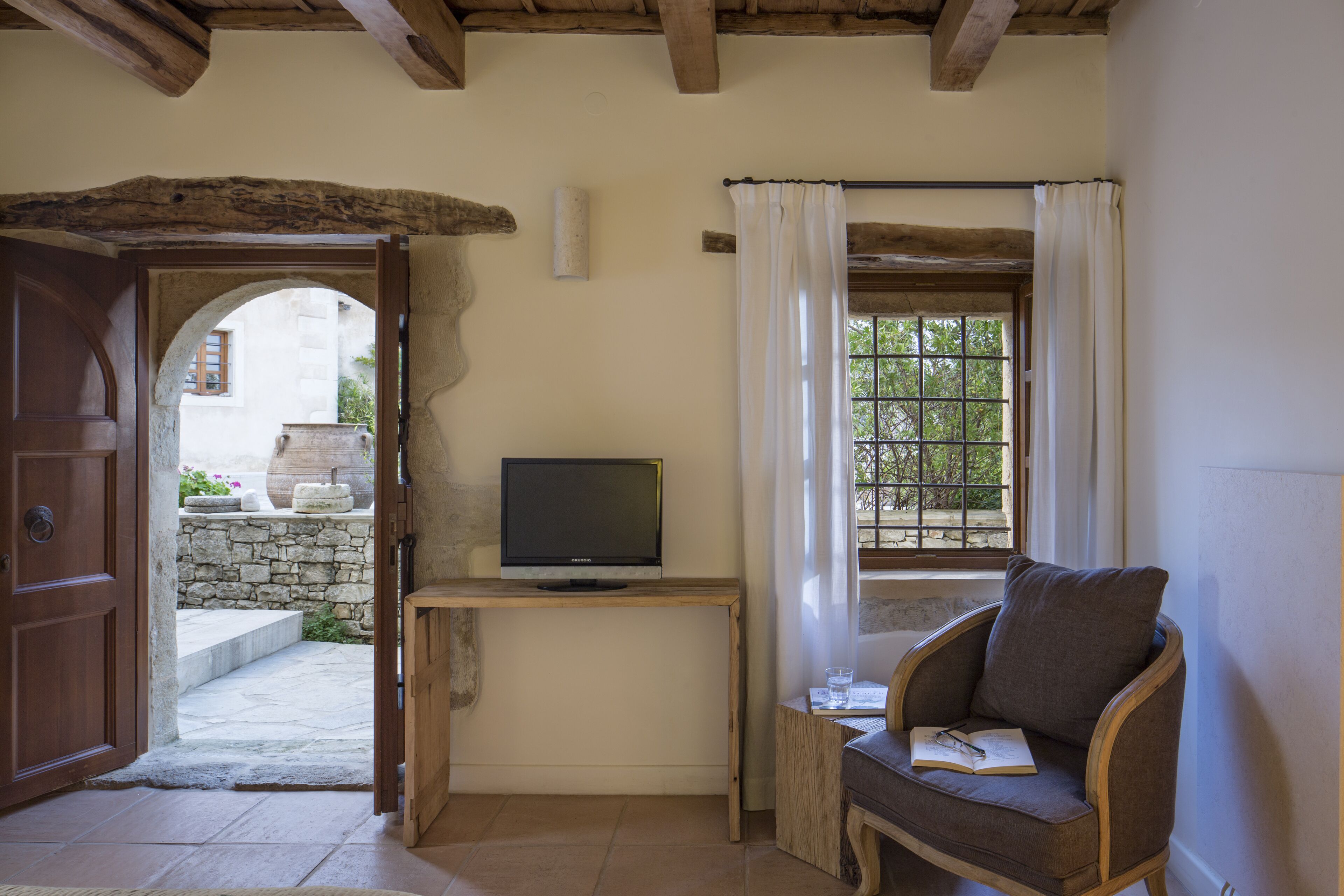 Quarto com parede de pedra, porta de madeira, televisão sobre suporte de madeira, janela com cortinas e cadeira com um livro.