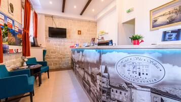 Um hall de hotel com paredes de pedra, uma mesa de recepção e um mural de um edifício histórico. Há cadeiras cor de verde-água e uma pequena mesa com um vaso de flores.