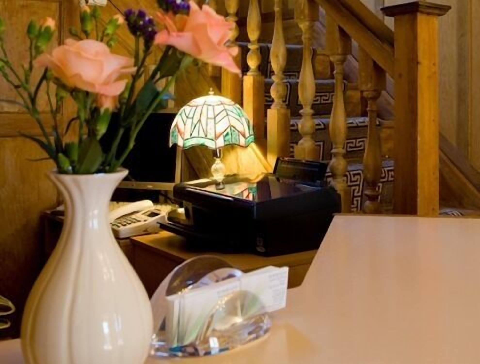Uma escada de madeira com guarda de padrão, um vaso branco com flores, uma mesa com abajur e um telefone.