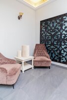 Quarto com duas poltronas confortáveis, uma pequena mesa com objetos decorativos, luminária fixada na parede e painel decorativo com padrão geométrico.