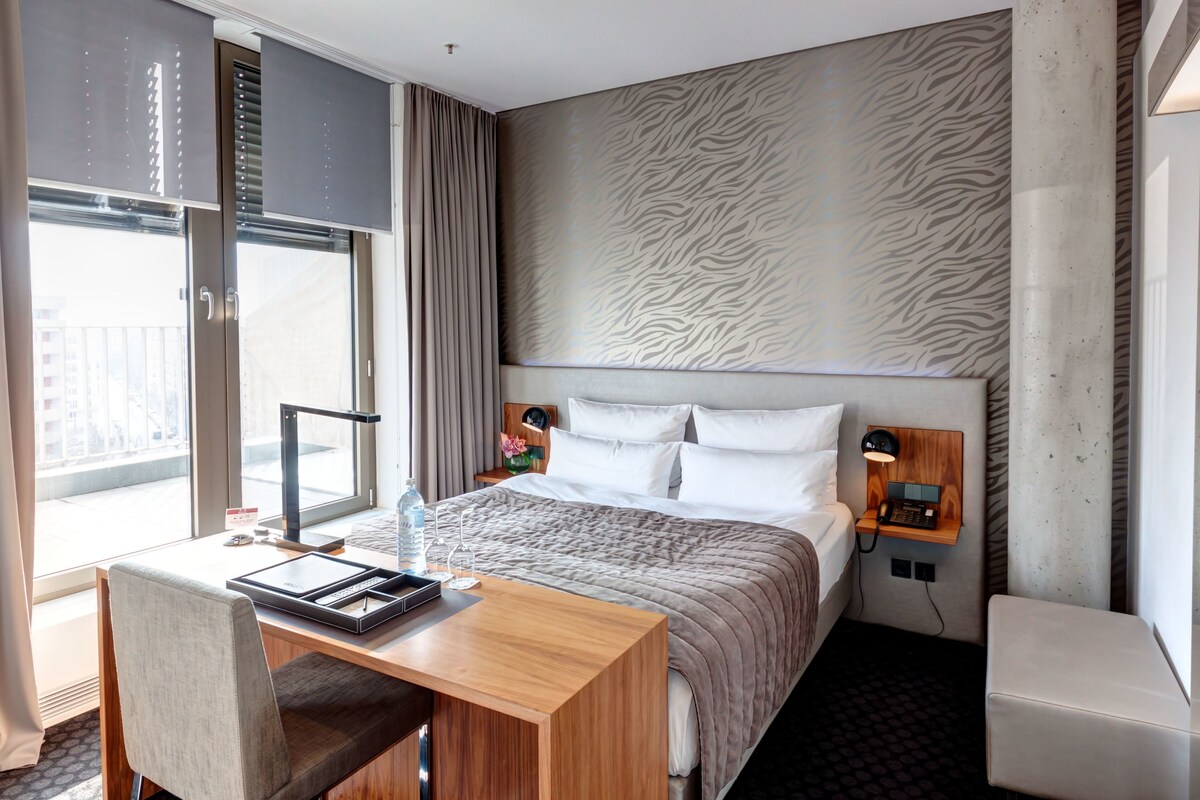 Quarto de hotel com cama, escrivaninha, cadeira e uma janela grande.