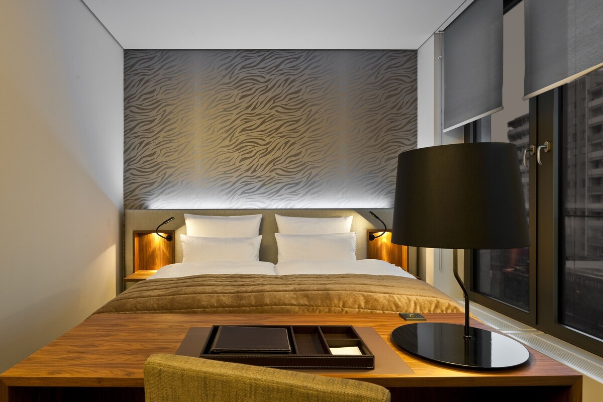 Quarto de hotel com cama, abajures, uma escrivaninha com cadeira, luminária e janela com vista.