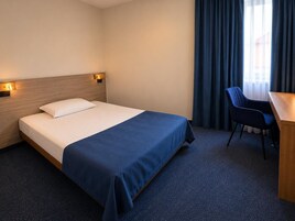 Quarto de hotel com uma cama, uma escrivaninha, uma cadeira e uma janela com cortinas.