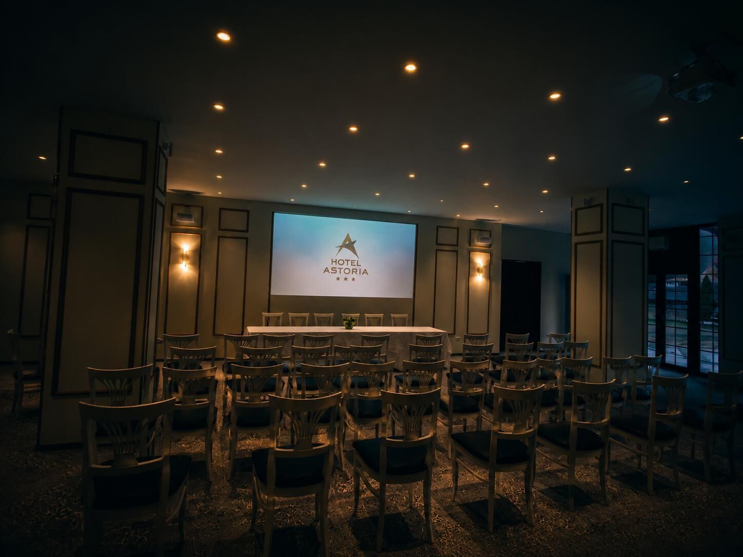 Uma sala de conferências com uma tela grande exibindo 'HOTEL ASTORIA' e uma classificação de três estrelas.