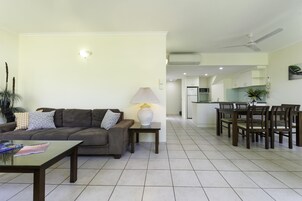 Sala de estar com sofá, mesa de centro e luminária, que dá acesso a uma área de jantar com mesa e cadeiras. Uma cozinha é visível ao fundo.