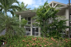 Um edifício com vários andares e área externa coberta, rodeado por vegetação exuberante e plantas floridas.