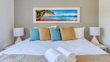 Cama bem arrumada com edredom branco e quatro travesseiros em cores variadas, uma cena de praia emoldurada na parede e duas luminárias de cabeceira.